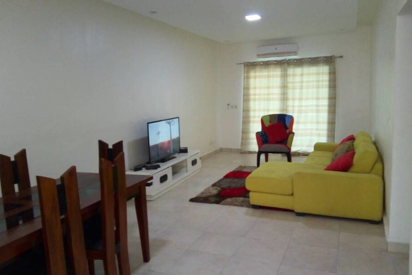 Uma sala de estar com um sofá amarelo, uma televisão em um móvel, uma mesa de jantar com cadeiras e uma poltrona colorida.