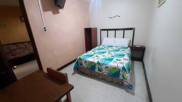 Quarto com cama, escrivaninha, cadeira, televisão na parede e uma pequena mesa.
