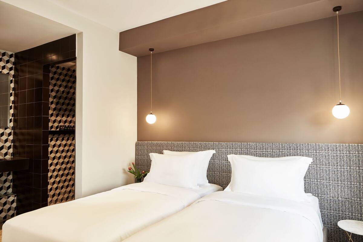 Quarto de hotel com design moderno, apresentando uma cama grande com roupa de cama branca, cabeceira texturizada e iluminação pendente.