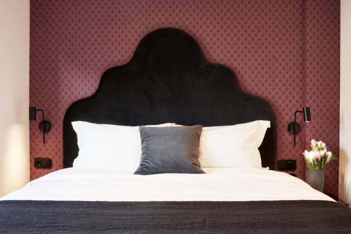 Uma cama com cabeceira escura, almofadas brancas e cinzentas e um edredom escuro.