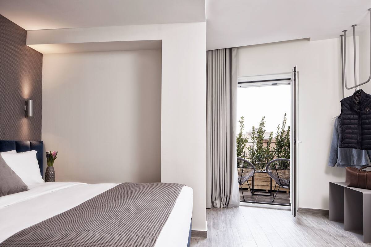 Quarto de hotel moderno com uma cama grande, varanda com área de estar externa e vista para árvores.