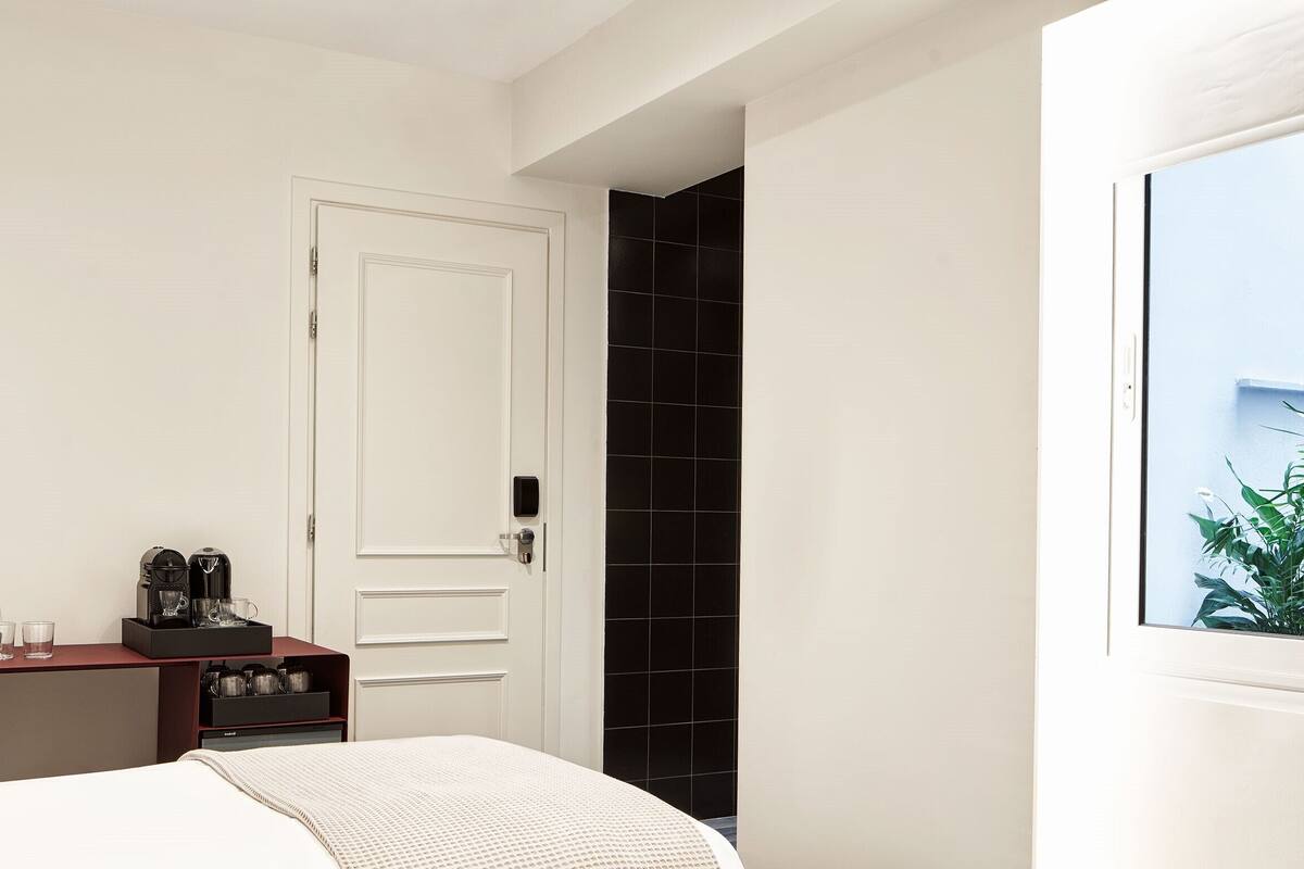 Quarto de hotel com cama, mesa de cabeceira com cafeteira, armário pequeno e porta para um banheiro com azulejos pretos.