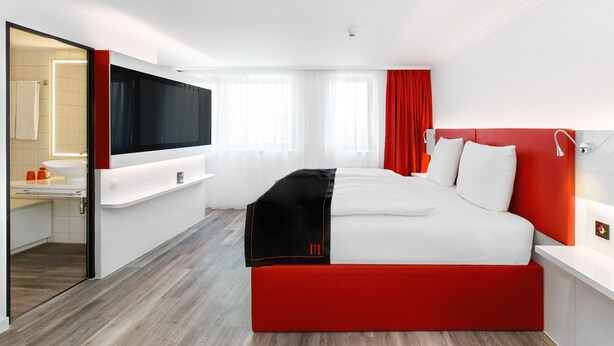 Quarto de hotel moderno com uma cama grande, televisão, parede de destaque vermelha e banheiro visível por uma porta aberta.