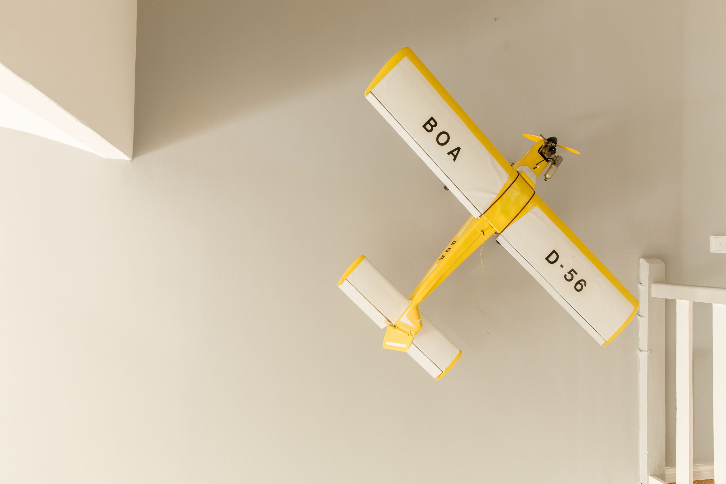 Um avião modelo com a inscrição "BOA D-56" nas asas, suspenso em um quarto com teto e corrimão brancos ao fundo.