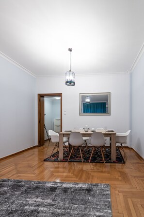 Uma sala de jantar com mesa de madeira, cadeiras brancas, cortina azul e luminária pendente.