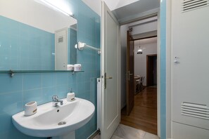 Casa de banho com lavatório branco, azulejos azuis, prateleira de vidro e porta que dá para um corredor.