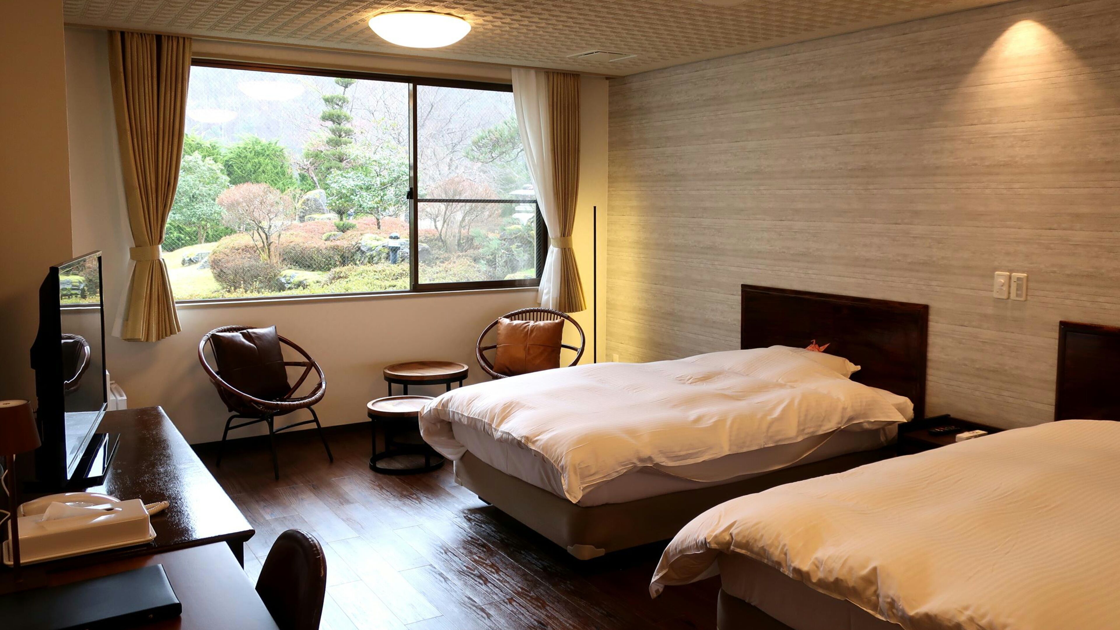 Quarto de hotel com duas camas, uma mesa de escritório, uma cadeira, uma mesinha e vista para um jardim pela janela.