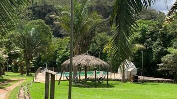 Paisagem verde exuberante com uma construção de telhado de palha, uma rede e área de piscina.