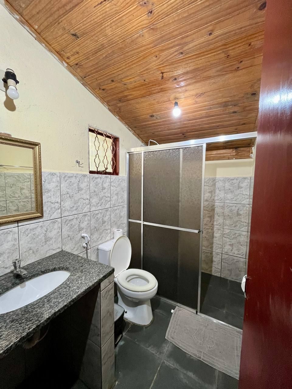 Casa de banho com bancada de granito, sanita, cabine de duche e teto de madeira.