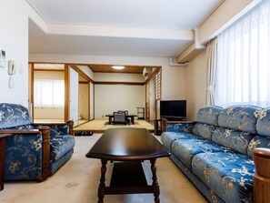 Uma sala de estar japonesa tradicional com tatames, uma mesa baixa, sofás estampados em azul e uma televisão.