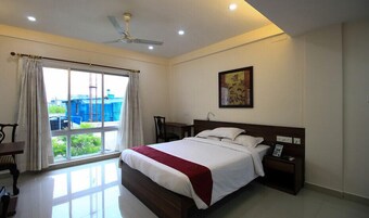 Quarto de hotel com cama, escrivaninha, cadeira, janela com cortinas, ventoinha no teto e um quadro na parede.