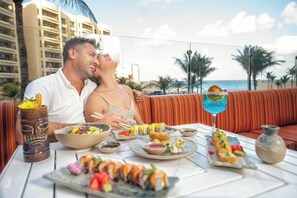 Um casal jantando em uma varanda com vista para a praia e palmeiras.