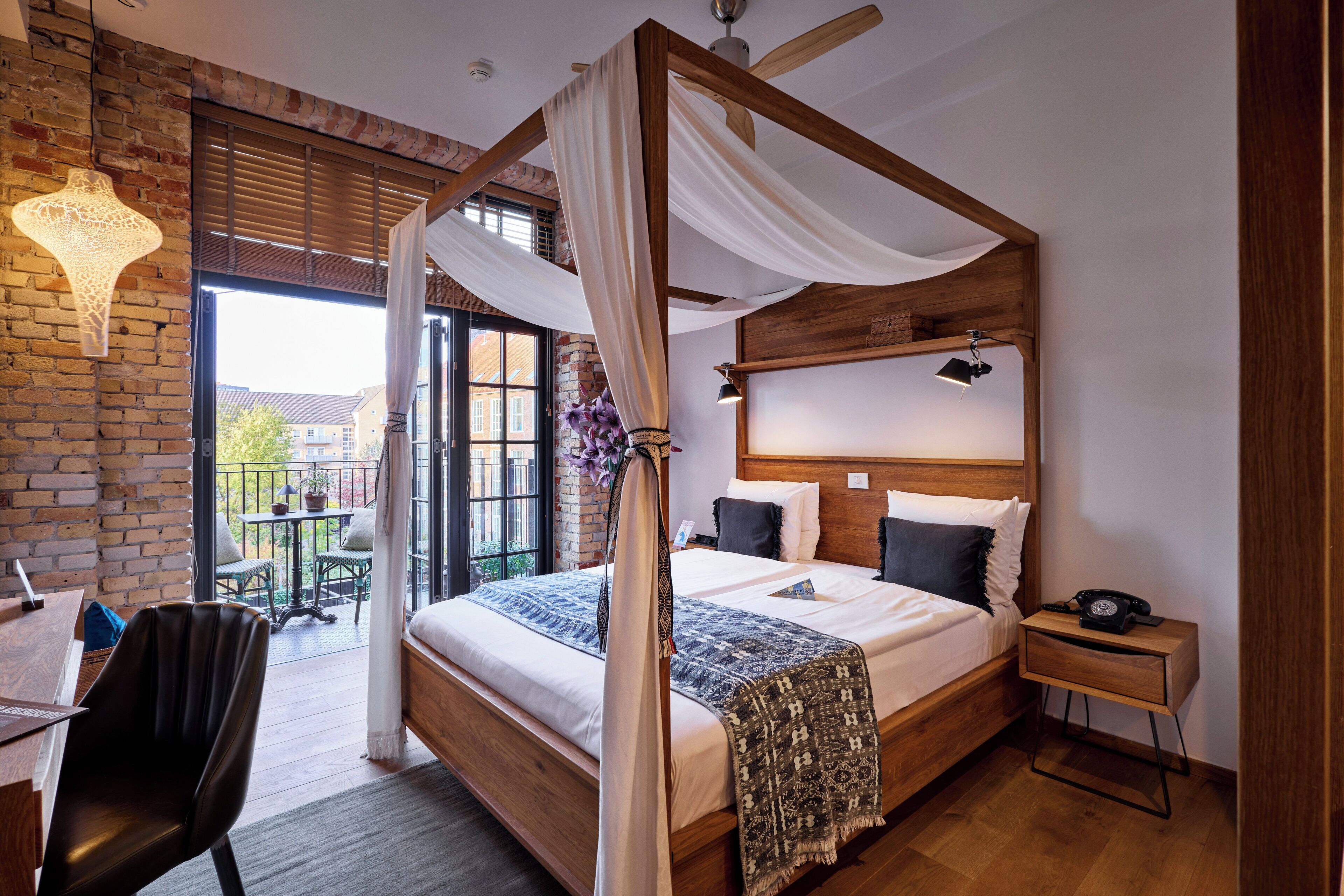 Uma cama com dossel e cortina, cabeceira de madeira, mesa de cabeceira com abajur e vista para o exterior através de uma porta de vidro.