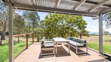 Um deck coberto ao ar livre com mesa de madeira e dois sofás, cercado por guarda-corpo de metal e com vista para uma área gramada com árvores.