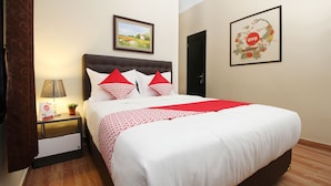 Uma cama bem arrumada com roupa de cama branca e vermelha, uma mesinha de cabeceira com abajur e livros, e duas molduras com quadros na parede.