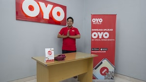 Uma pessoa em frente a um balcão com uma faixa vermelha da OYO Hotels e uma grande placa OYO na parede.
