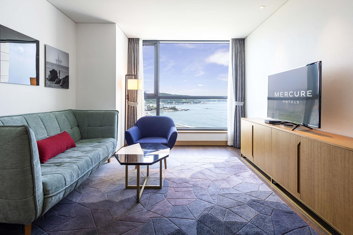 Quarto de hotel com uma janela grande, um sofá verde, uma poltrona azul, uma mesa pequena e uma televisão sobre um móvel de madeira.