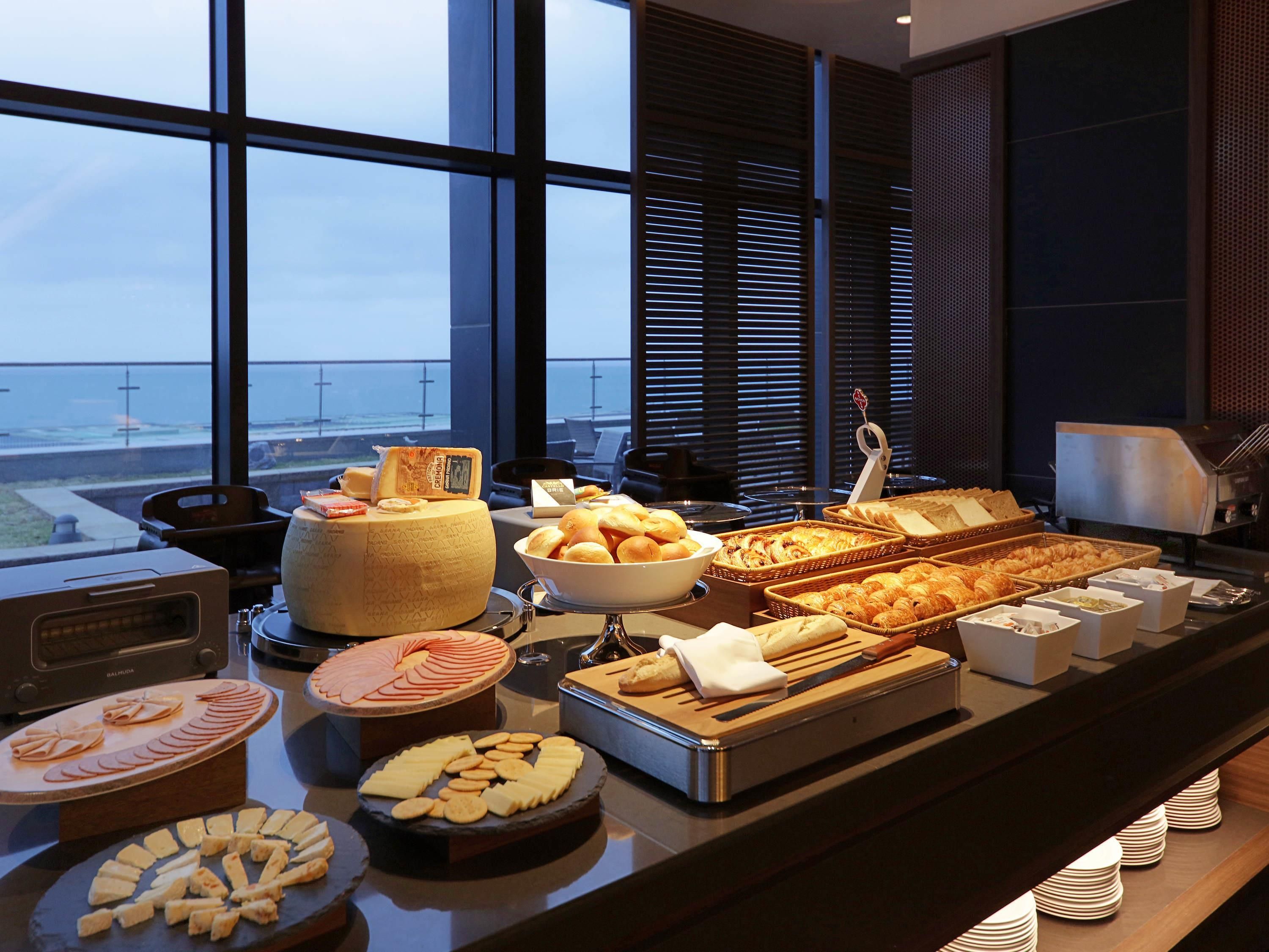 Buffet variado com queijos, pães e pastéis, com vista para o oceano através de grandes janelas.