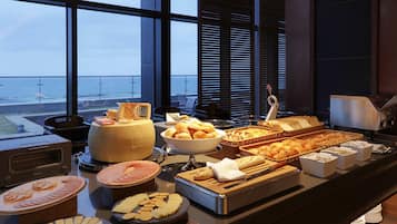 Buffet variado com queijos, pães e pastéis, com vista para o oceano através de grandes janelas.