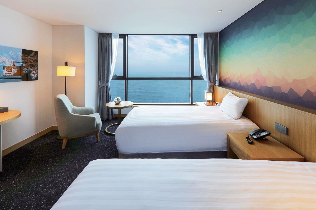 Quarto de hotel com duas camas, uma mesa, uma cadeira, uma mesinha, uma luminária, um telefone e uma janela grande com vista para o mar.