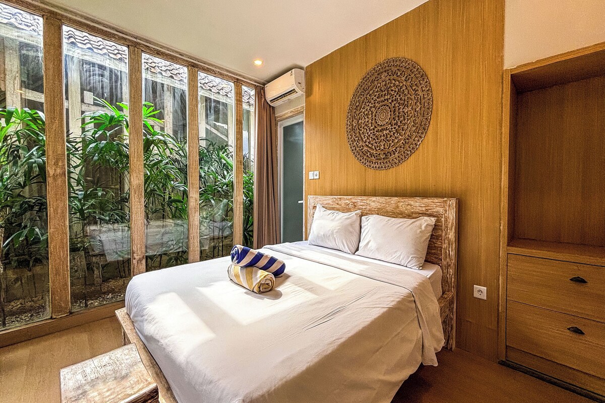 Quarto com uma cama grande, cabeceira de madeira, um objeto decorativo na parede e vista para o verde através de janelas amplas.
