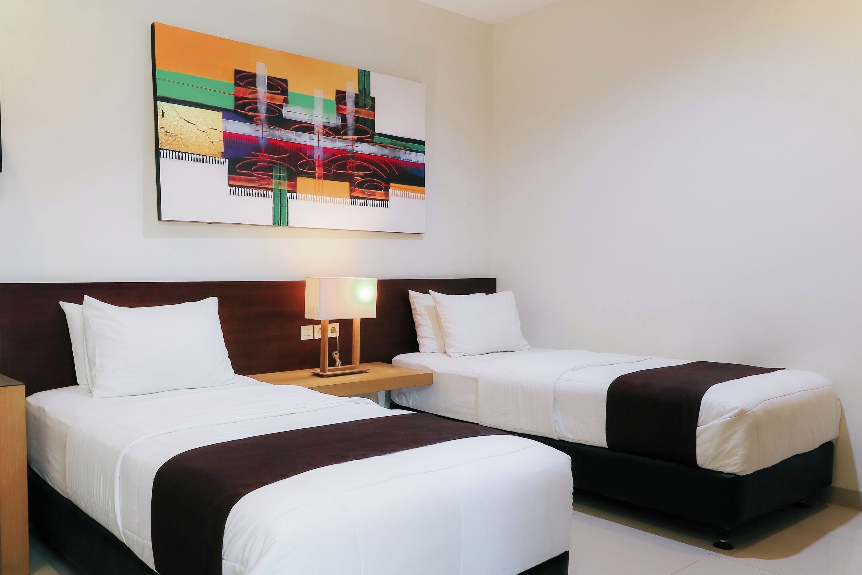 Quarto de hotel com duas camas, uma mesa de cabeceira com abajur e um quadro abstrato colorido na parede.