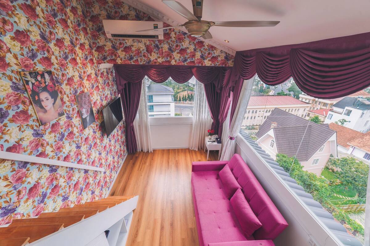 Quarto com piso de madeira, sofá rosa, ventilador de teto e janela com cortinas.