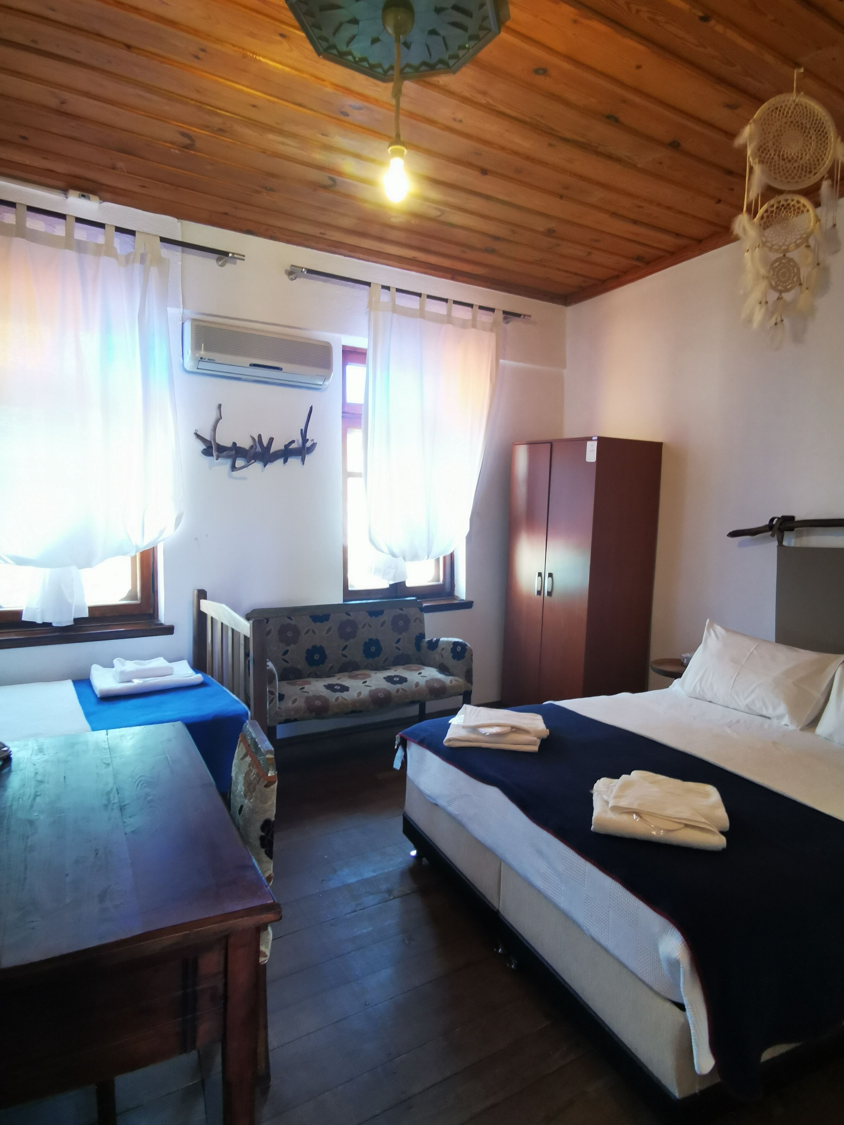Um quarto aconchegante com teto de madeira, cama com colcha azul, sofá com tecido floral, mesa de madeira e um guarda-roupa.