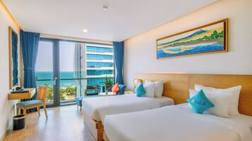 Quarto de hotel com duas camas, uma escrivaninha, uma cadeira e vista para o oceano.