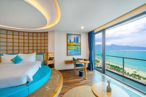 Quarto de hotel com cama, escrivaninha, cadeira, vista para a praia e um quadro na parede.