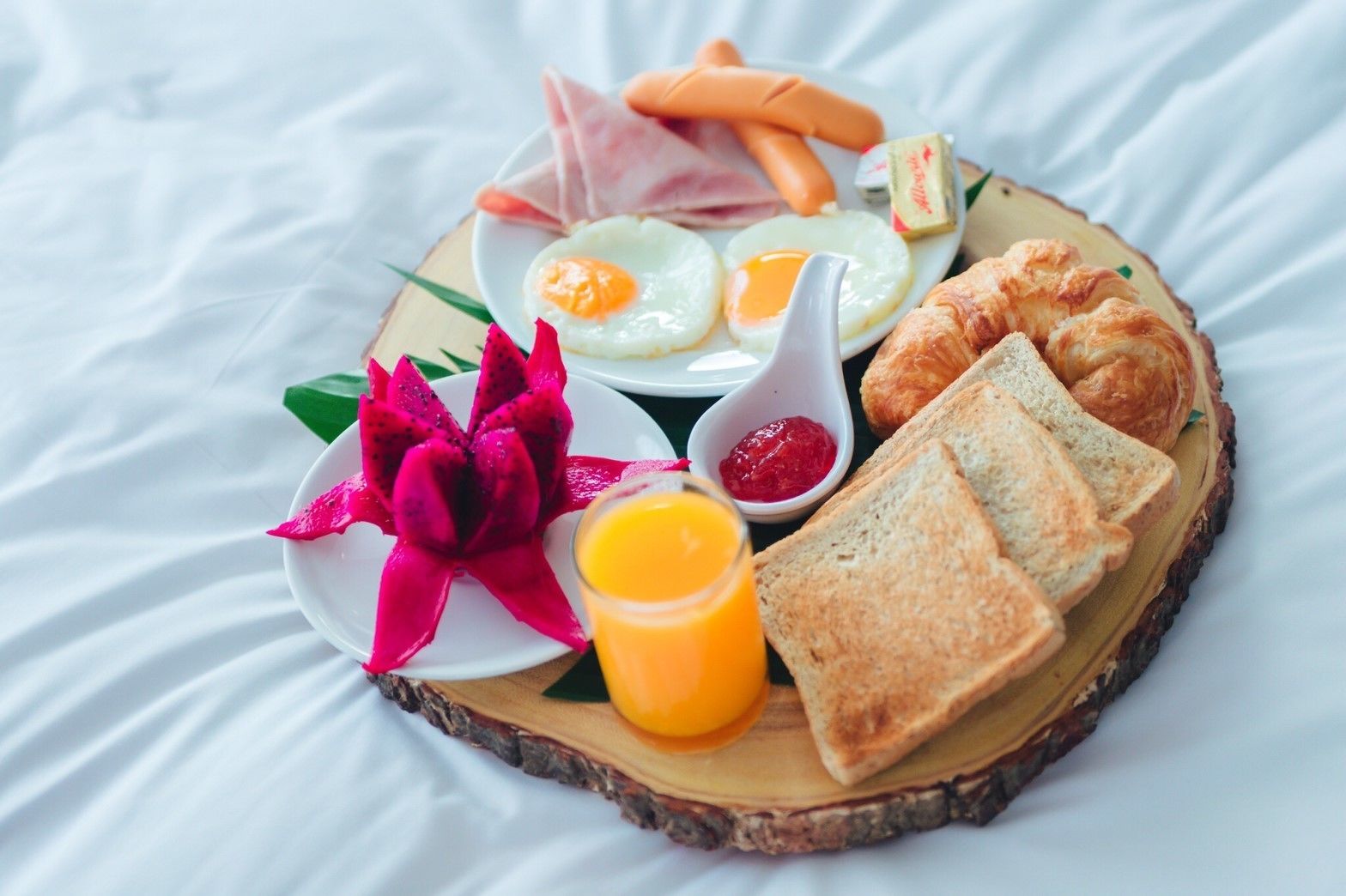 Bandeja de café da manhã com ovos, salsichas, croissants, torradas e uma decoração de frutas sobre uma tábua de madeira.