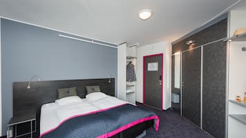 Quarto de hotel moderno com cama, mesa de cabeceira, guarda-roupa e um pequeno estante com artigos de higiene pessoal.