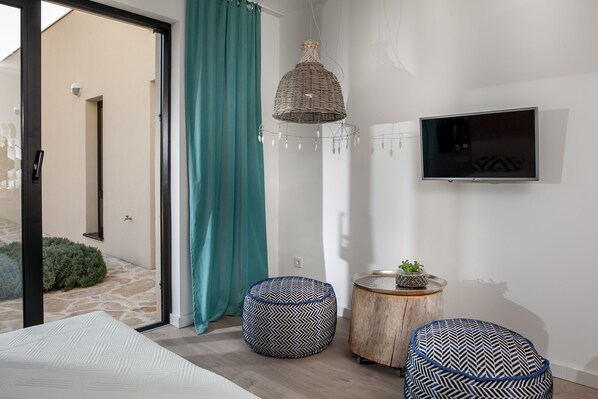 Sala moderna com uma televisão grande, dois pufes estampados, uma luminária pendente e vista para um jardim através de uma porta aberta.