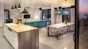 Cozinha moderna com ilha central, área de jantar com cadeiras transparentes e vista para a cidade através de grandes janelas.