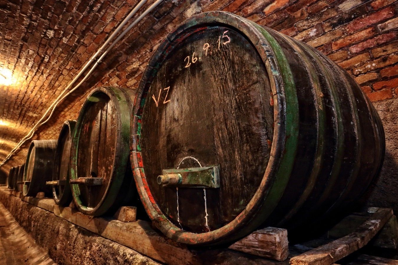 Barricas de madeira alinhadas em uma adega de paredes de tijolo com pouca luz.