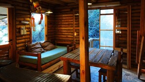 Interior de uma cabana de tronco com um banco de madeira, uma mesa e uma janela com vista para árvores.