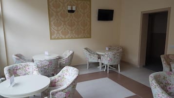 Quarto com cadeiras e mesas de padrão floral, uma TV de tela plana na parede e uma porta que dá para outra área.