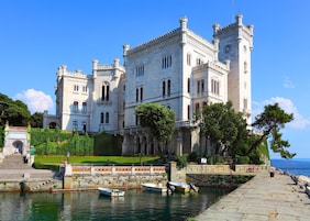 Um edifício grande e histórico com uma torre imponente e janelas em arco, localizado à beira-mar, com barcos atracados.