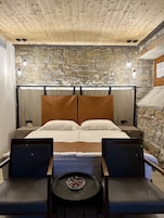 Quarto moderno com teto de madeira, parede de pedra, uma cama de casal com cabeceira marrom, duas mesas de cabeceira, uma pequena mesa redonda com cadeiras e luminárias pendentes.