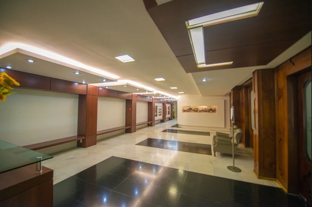 Um hall de hotel com design moderno, destacando-se um balcão de recepção em vidro, uma área de estar e um corredor que dá para outros quartos.