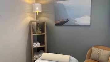 Quarto com cama, uma cadeira, prateleira com toalhas, estátua de Buda, abajur e pintura de uma paisagem montanhosa.