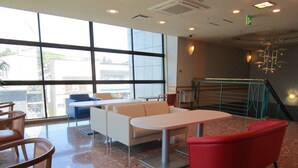 Um espaço interior moderno com área de estar, incluindo um sofá branco, uma cadeira vermelha e uma mesa. Grandes janelas proporcionam luz natural e uma vista para o exterior.