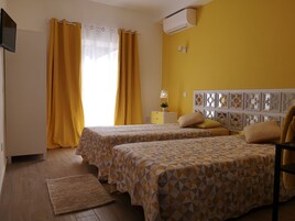 Quarto de hotel com duas camas, uma pequena mesa de cabeceira com abajur, ar condicionado fixado na parede e cabeceira decorativa.