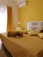 Quarto com decoração amarela, uma cama, um vaso de planta e ar condicionado fixado na parede.