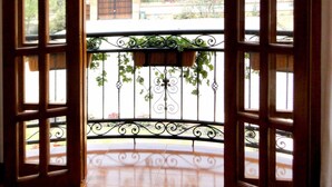 Uma porta de madeira aberta com vista para uma varanda com grade de metal e plantas penduradas.