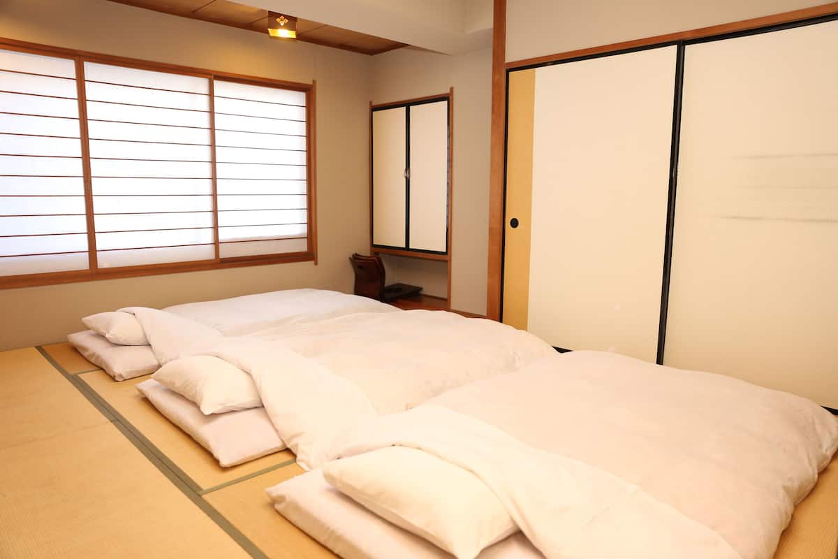 Um quarto japonês tradicional com piso de tatame, uma cama grande com roupa de cama branca, uma porta de correr e uma janela com biombos shoji.