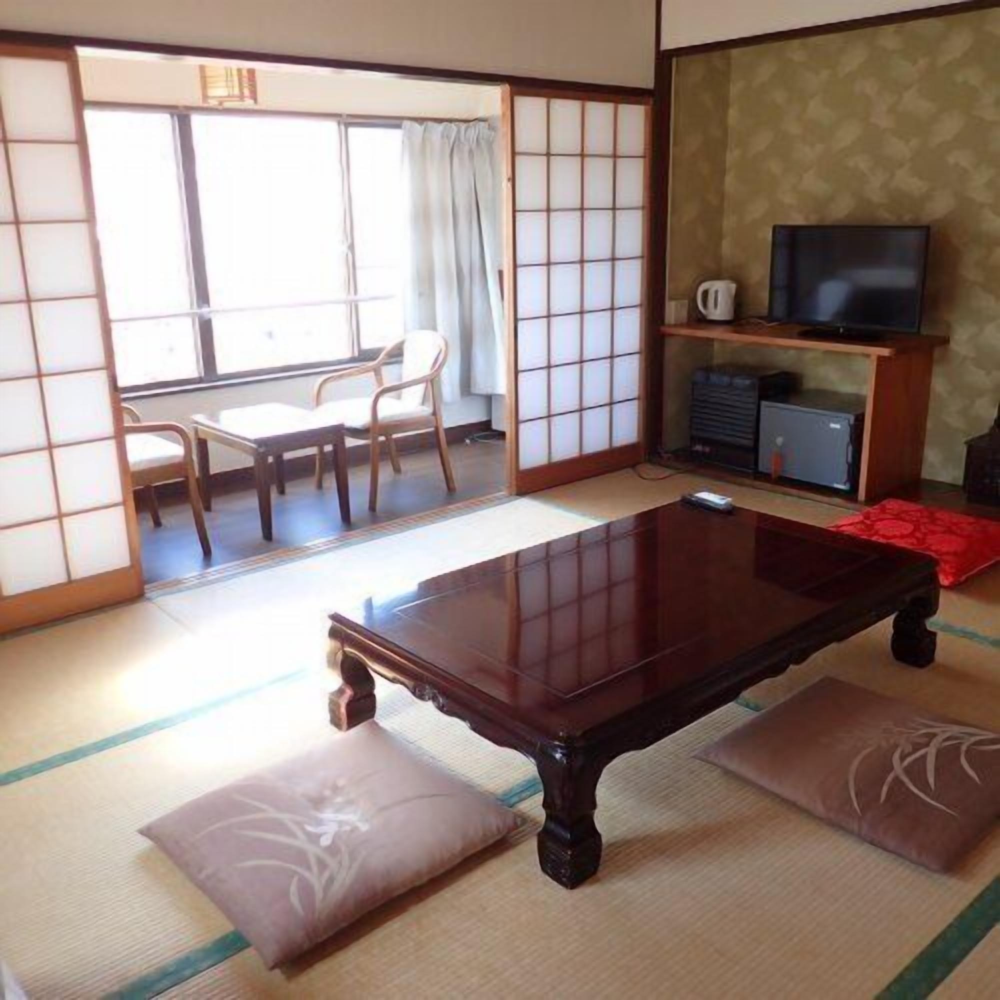 Um quarto tradicional japonês com tatames, uma mesa de madeira baixa, uma televisão em suporte e uma porta deslizante que dá para uma varanda.
