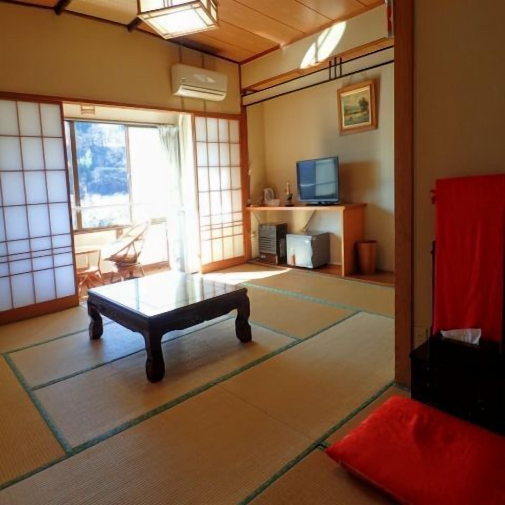 Quarto japonês tradicional com tatames, uma mesa baixa, uma televisão e uma porta deslizante para uma área externa.