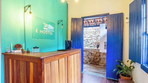 Uma receção com acabamento em madeira, uma placa com a inscrição "É HOSTEL" e uma parede de pedra visível através de uma porta aberta.