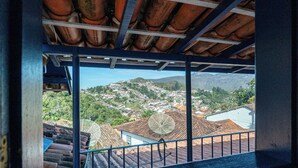 Vista de uma varanda com telhado de azulejos e grades de metal, com vista para uma cidade com casas e colinas.